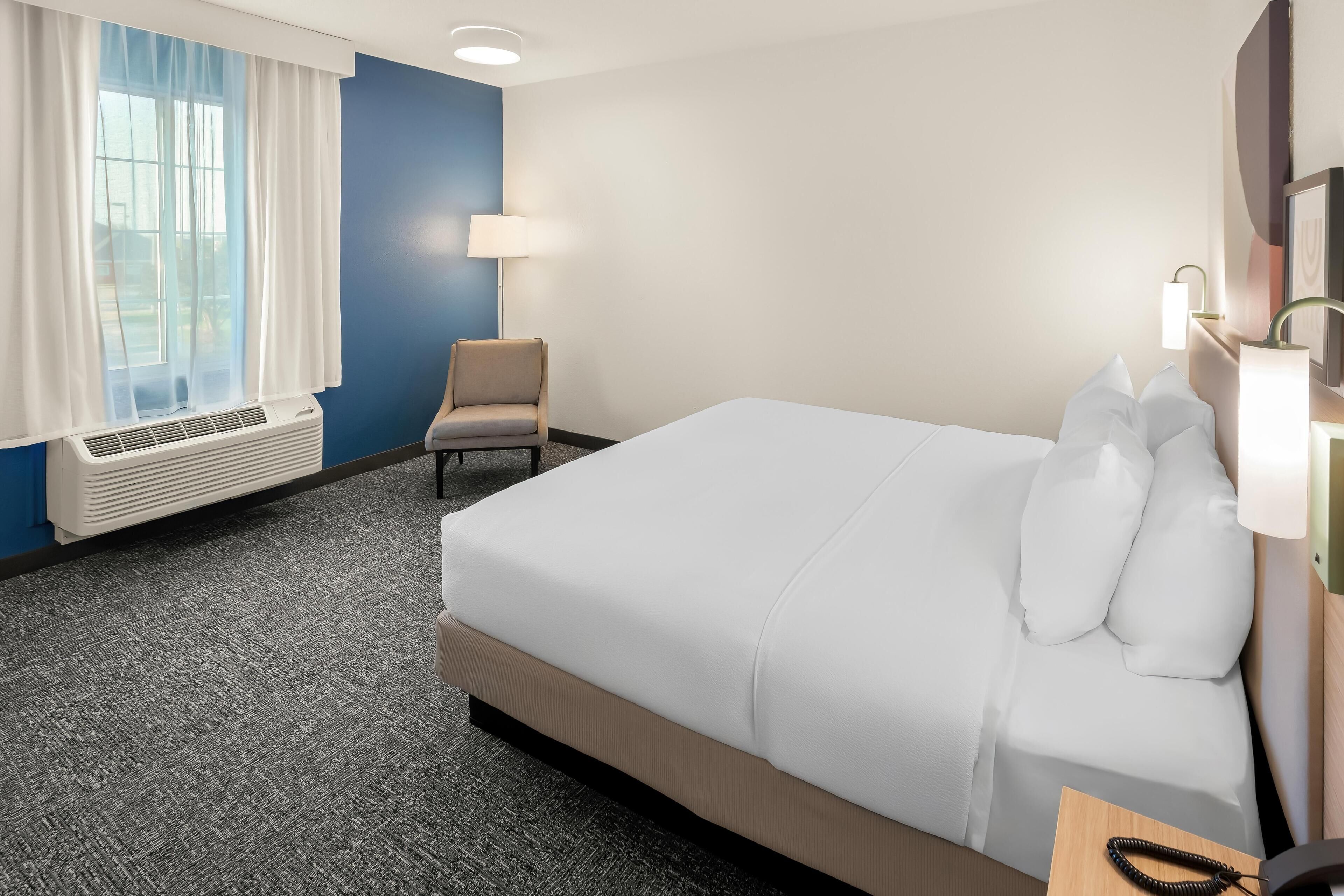 Quarto de hotel com cama, uma cadeira, escrivaninha com telefone, luminária e janela com cortinas.