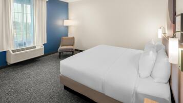 Quarto de hotel com cama, uma cadeira, escrivaninha com telefone, luminária e janela com cortinas.