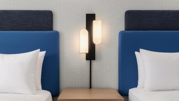 Quarto de hotel com duas camas, uma mesa de cabeceira e um candeeiro fixo na parede.