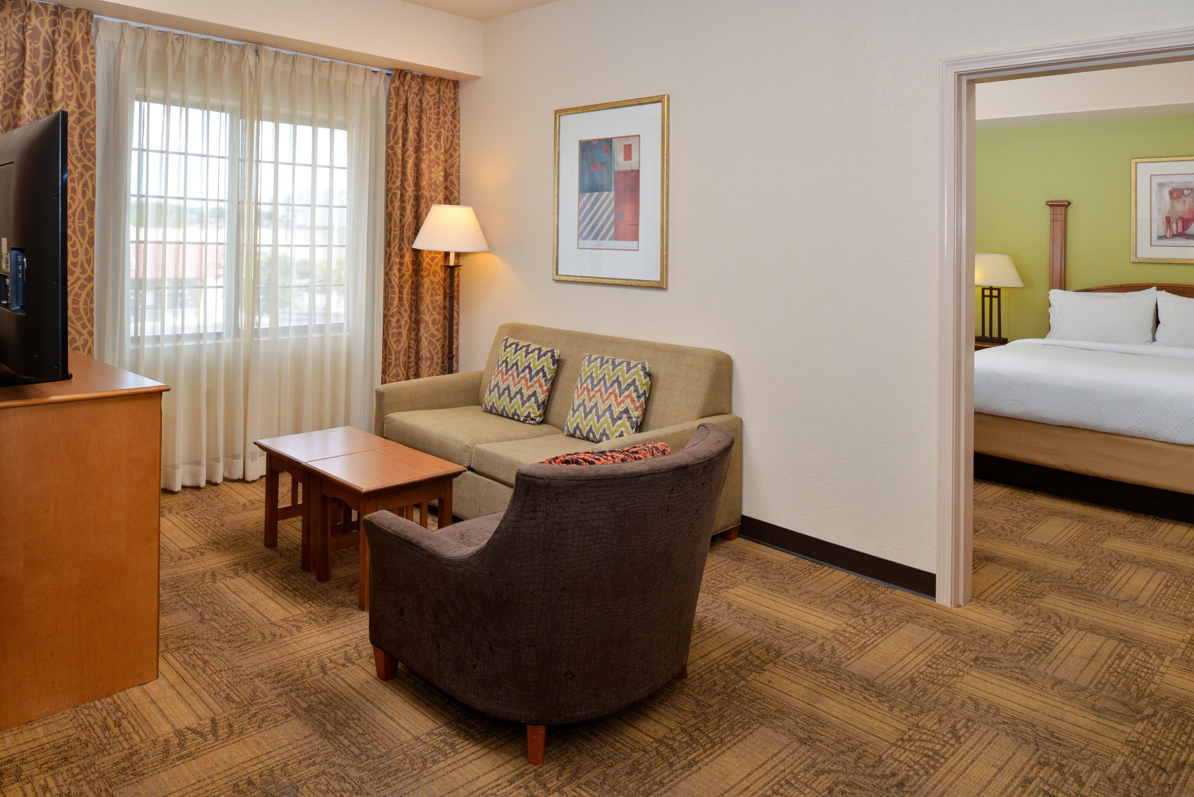 Quarto de hotel com sofá, poltrona, mesa de cabeceira, luminária, obra de arte emoldurada e televisão.