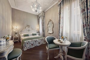 Quarto de hotel clássico com cama, duas cadeiras, uma mesinha, espelho e lustre.