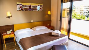 Quarto de hotel com uma cama grande, uma mesa de cabeceira com telefone, um criado-mudo, uma janela com vista para edifícios e uma obra de arte na parede.