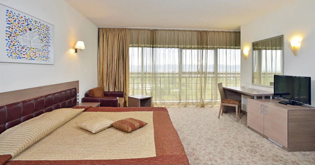 Quarto de hotel com uma cama grande, uma escrivaninha, televisão, cadeira e janela com cortinas.