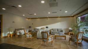 Uma sala de estar ampla, com diversas opções de assento, incluindo sofás e cadeiras, dispostas em torno de uma mesa de centro. O espaço possui uma grande janela que oferece luz natural e vista para o exterior.
