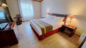 Quarto de hotel com uma cama, mesas de cabeceira, escrivaninha e cadeira, janela com cortinas e radiador.