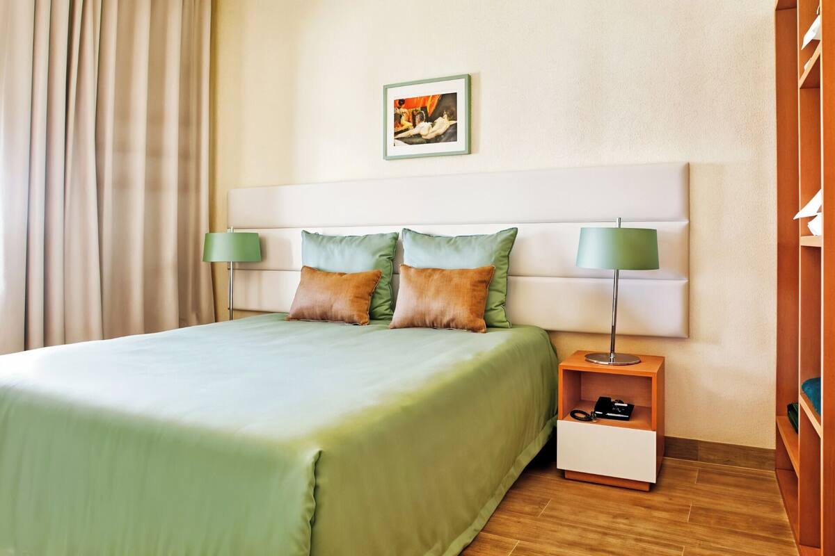 Uma cama bem arrumada com um cobre-leito verde, dois travesseiros, uma mesa de cabeceira com uma luminária e uma pequena gaveta.