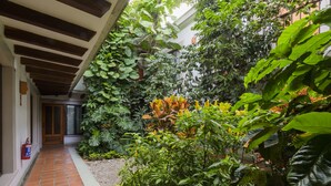 Uma galeria coberta com chão de azulejos, rodeada por vegetação exuberante e diversas plantas.