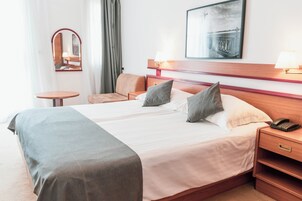 Quarto de hotel com uma cama grande, mesas de cabeceira, uma cadeira, um espelho e um quadro na parede.