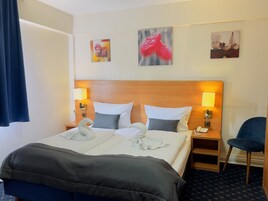 Quarto de hotel com uma cama grande, duas mesas de cabeceira com luminárias, uma poltrona e obras de arte nas paredes.