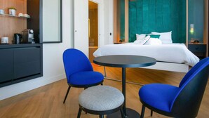 Quarto de hotel moderno com cama, duas cadeiras azuis, uma mesa redonda pequena, máquina de café e armário de madeira.