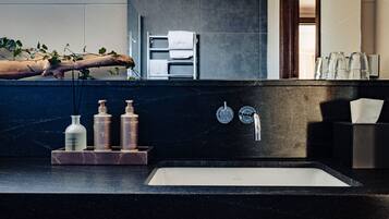 Casa de banho moderna com bancada de mármore preto, lavatório branco e um espelho que reflete um porta-toalhas e uma planta.