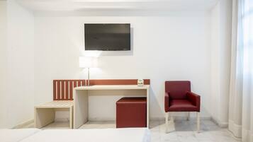Quarto de hotel com uma cama, uma escrivaninha com cadeira, televisão e luminária.