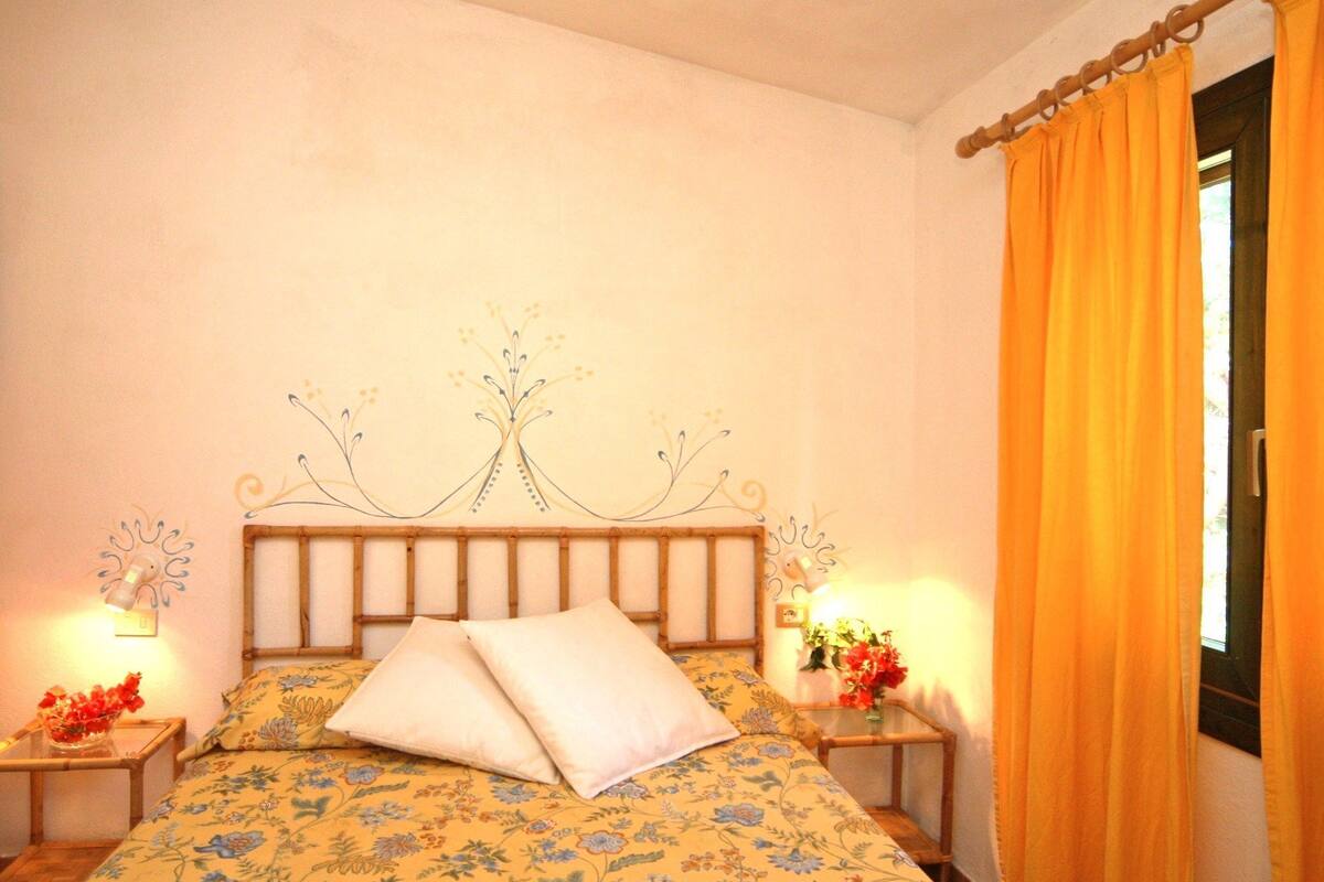 Uma cama com colcha florida, duas almofadas, cabeceira de madeira e duas mesas de cabeceira com abajures.