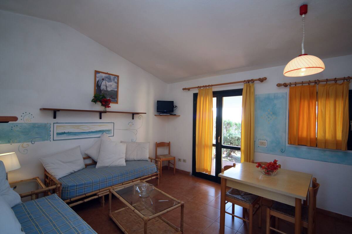 Quarto com sofá, mesa pequena, televisão e janela com cortinas.