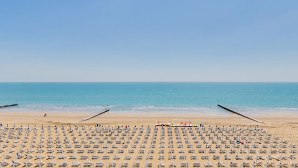Uma praia com fileiras de espreguiçadeiras e guarda-sóis, um píer e águas azuis calmas.