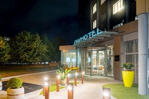Entrada de hotel com fachada de vidro, área de estar externa e espaço de acesso bem iluminado.