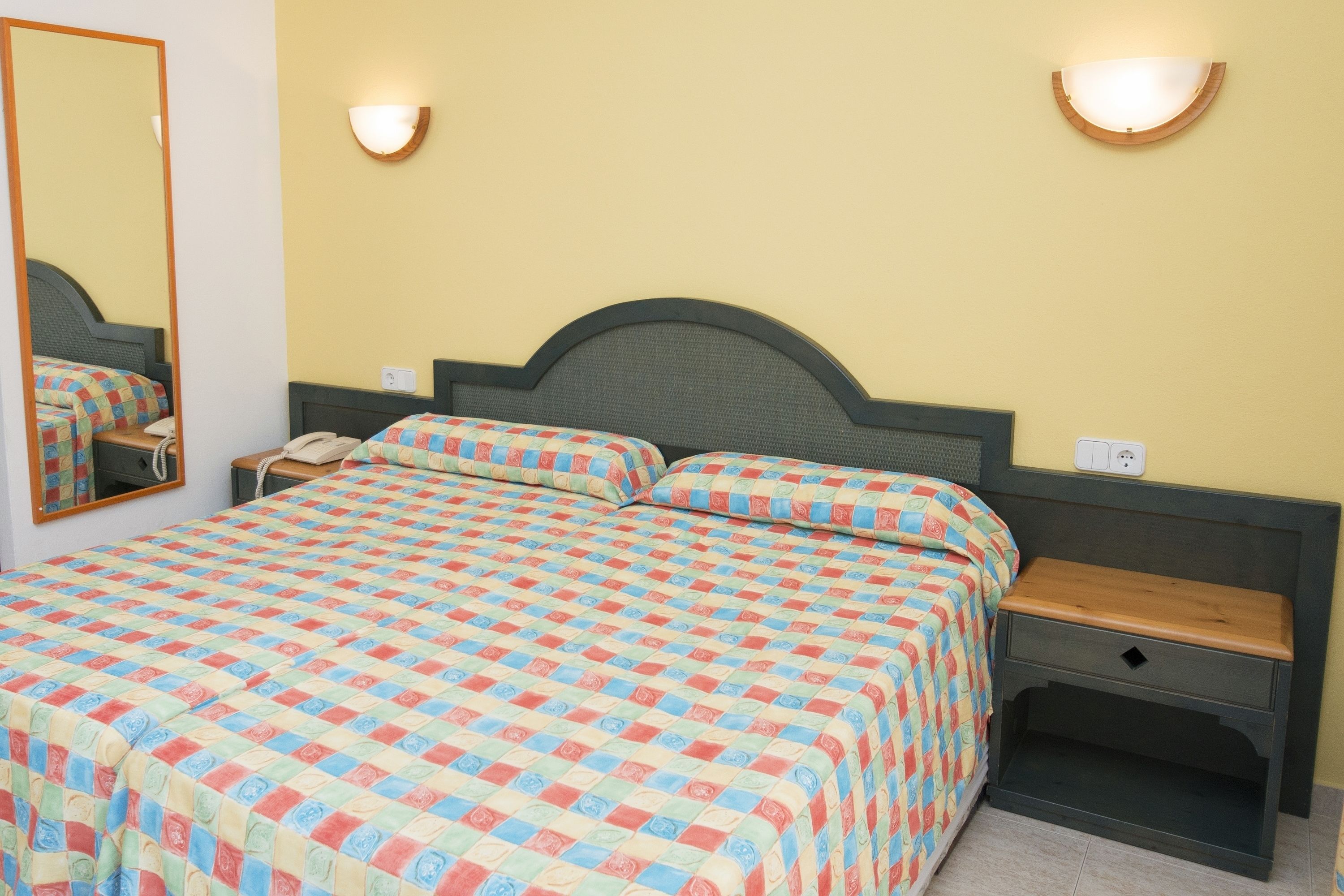 Quarto com uma cama grande, duas mesas de cabeceira, um criado-mudo, um espelho e duas luminárias fixadas na parede.