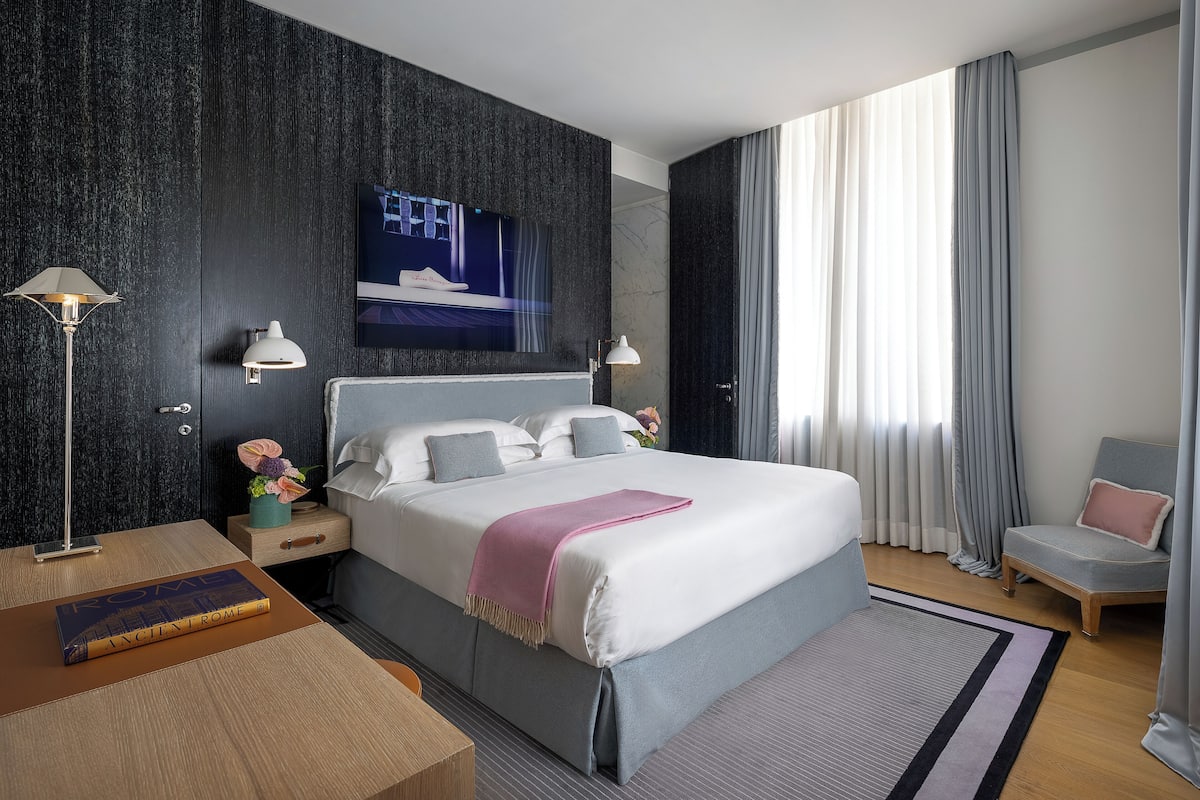 Quarto de hotel moderno com uma cama grande, televisão de tela plana, mesa de cabeceira com luminárias, uma cadeira e vista para o exterior.