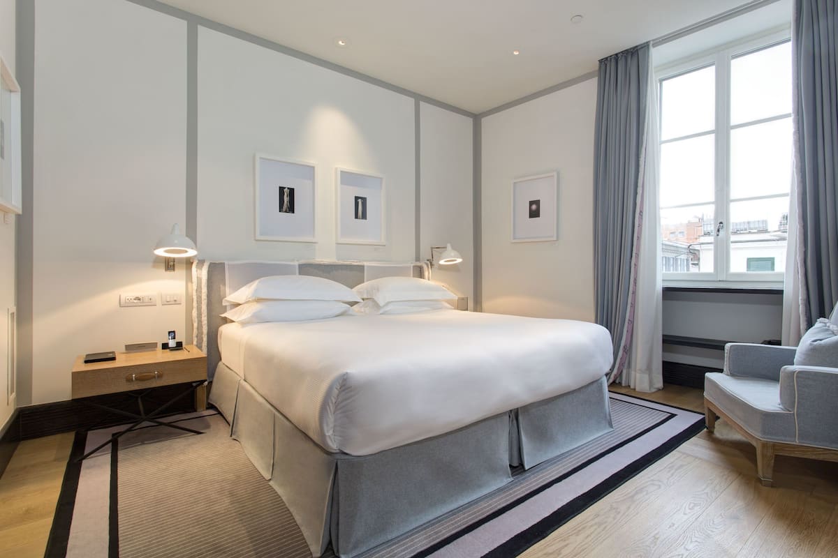 Quarto de hotel com uma cama grande, mesas de cabeceira, escrivaninha e uma cadeira. Existem quadros nas paredes e uma janela com cortinas.