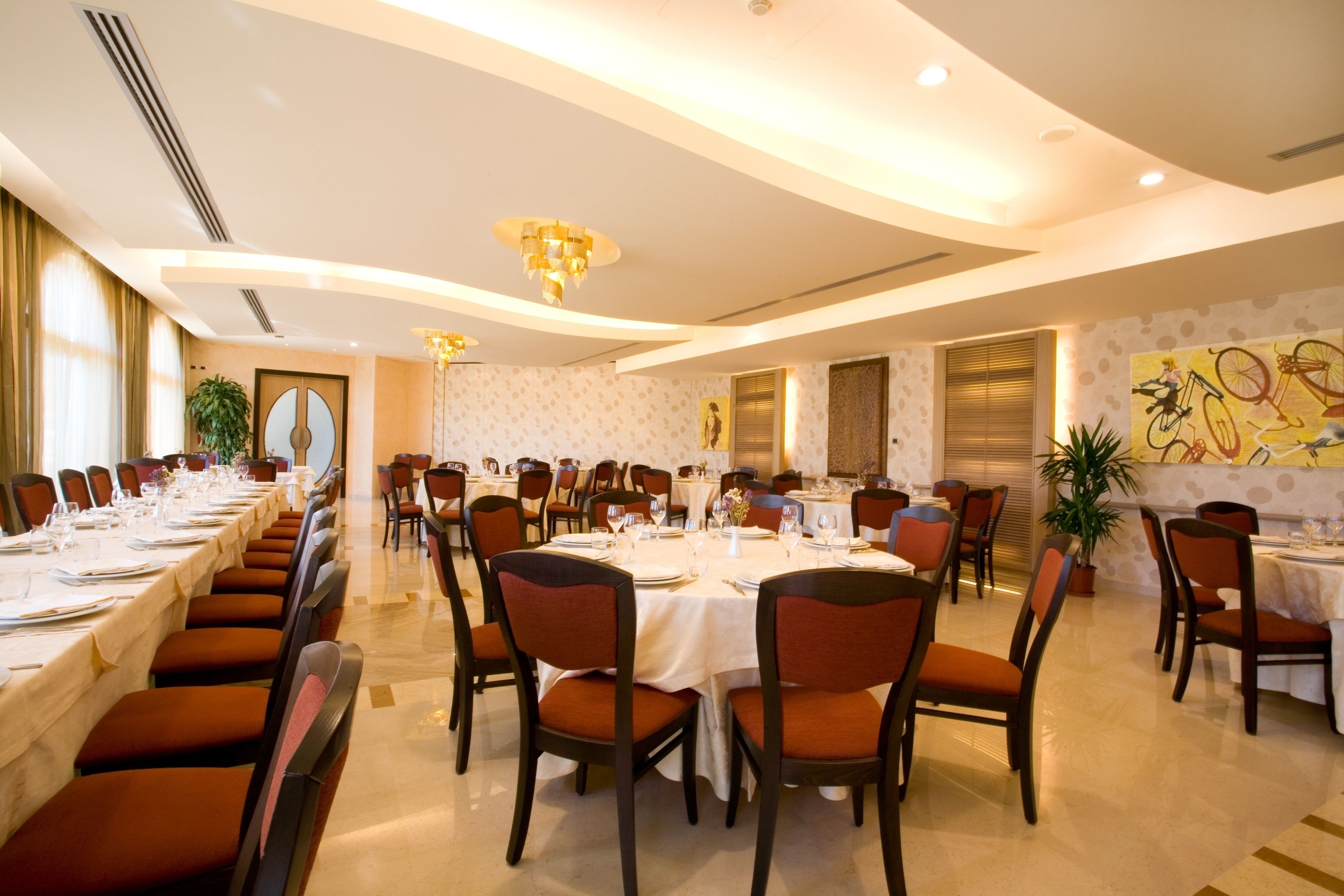 Uma sala de jantar ampla, com mesas compridas preparadas para vários hóspedes, rodeada por cadeiras.