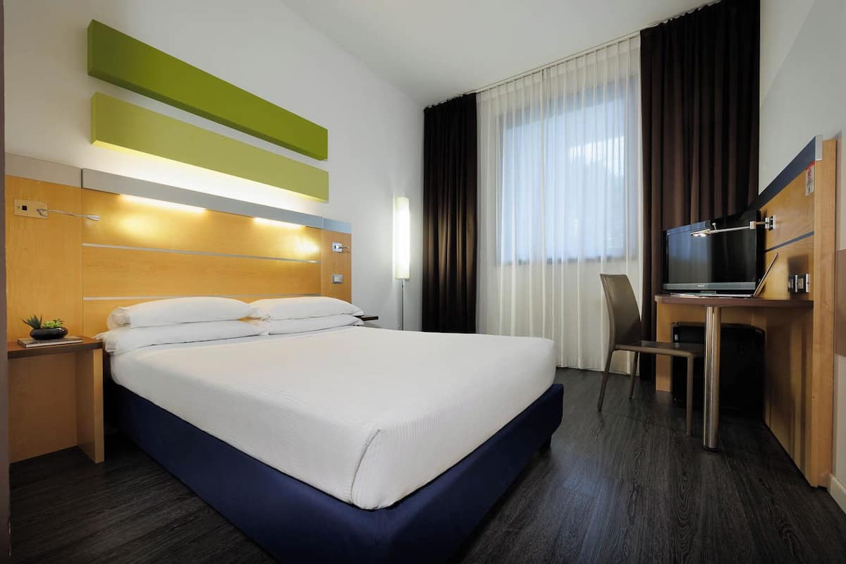 Quarto de hotel com uma cama grande, uma escrivaninha com televisão e uma janela com cortinas.