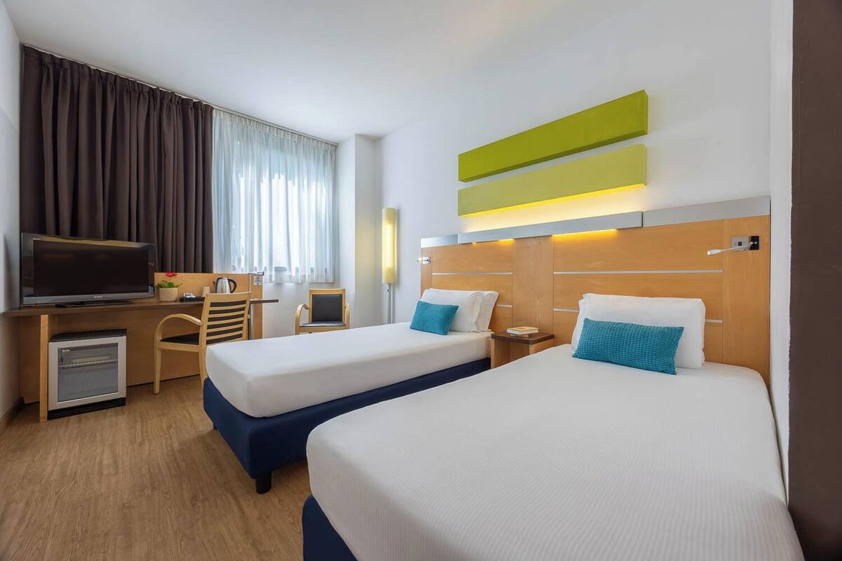 Quarto de hotel com duas camas, uma escrivaninha com televisão, um mini-frigobar e uma cadeira.