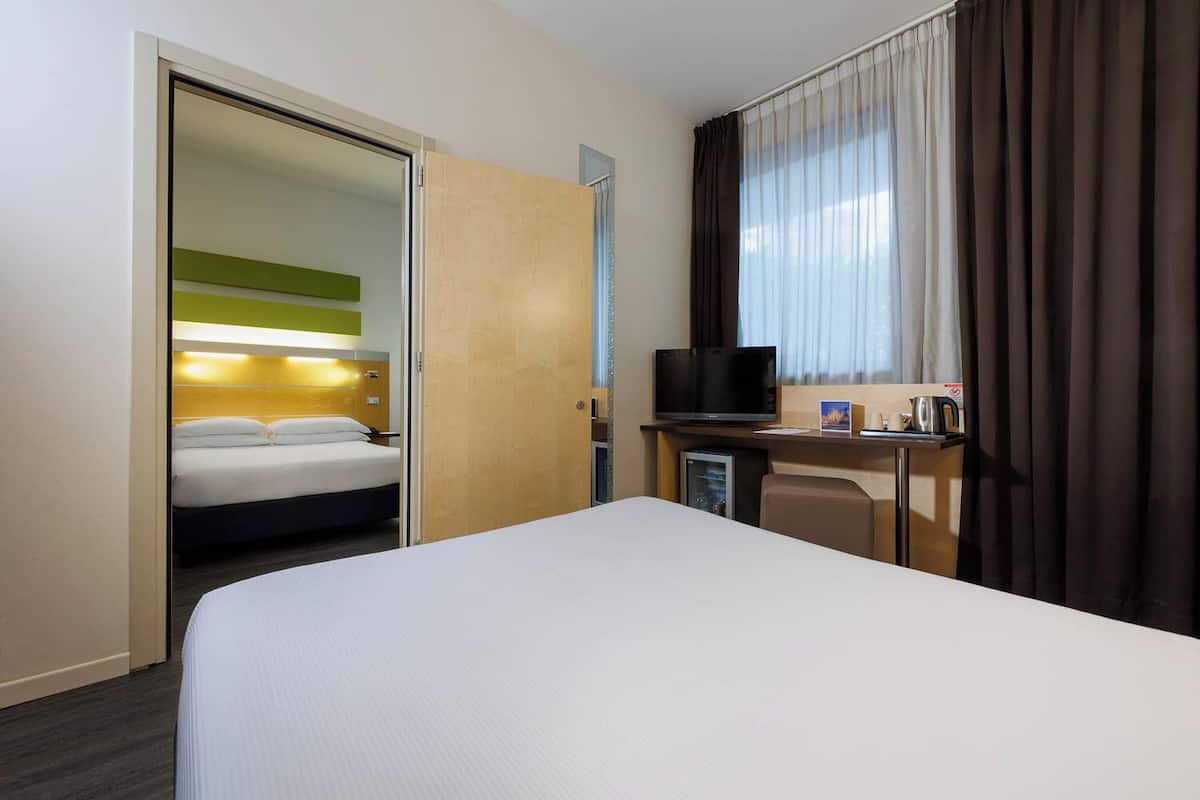 Quarto de hotel com cama, escrivaninha, televisão, mini-geladeira e janela com cortinas.