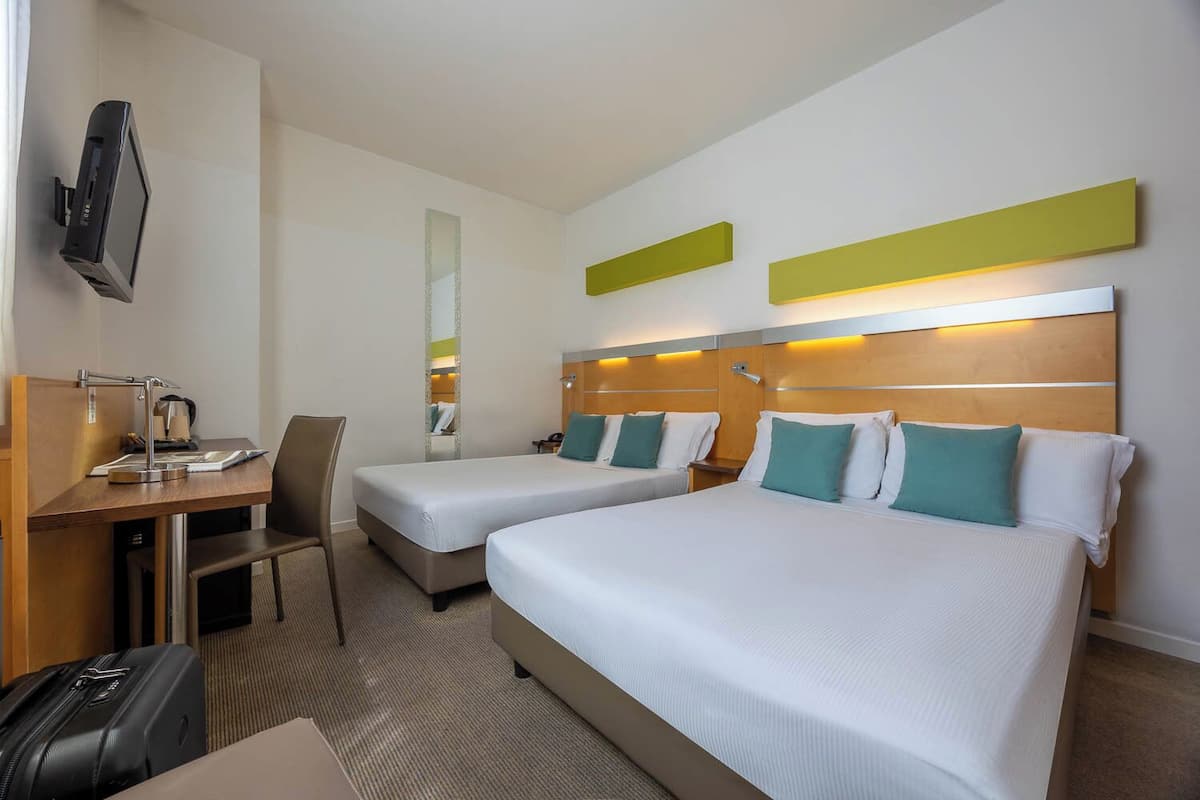 Quarto de hotel com duas camas, uma escrivaninha, uma cadeira, uma televisão e uma mala.