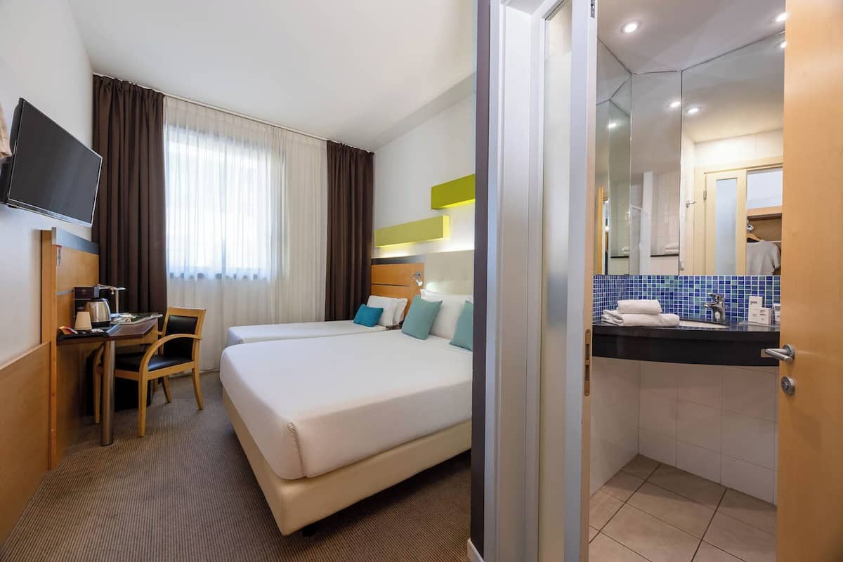 Quarto de hotel com cama, escrivaninha com cadeira, televisão e banheiro com espelho e pia.
