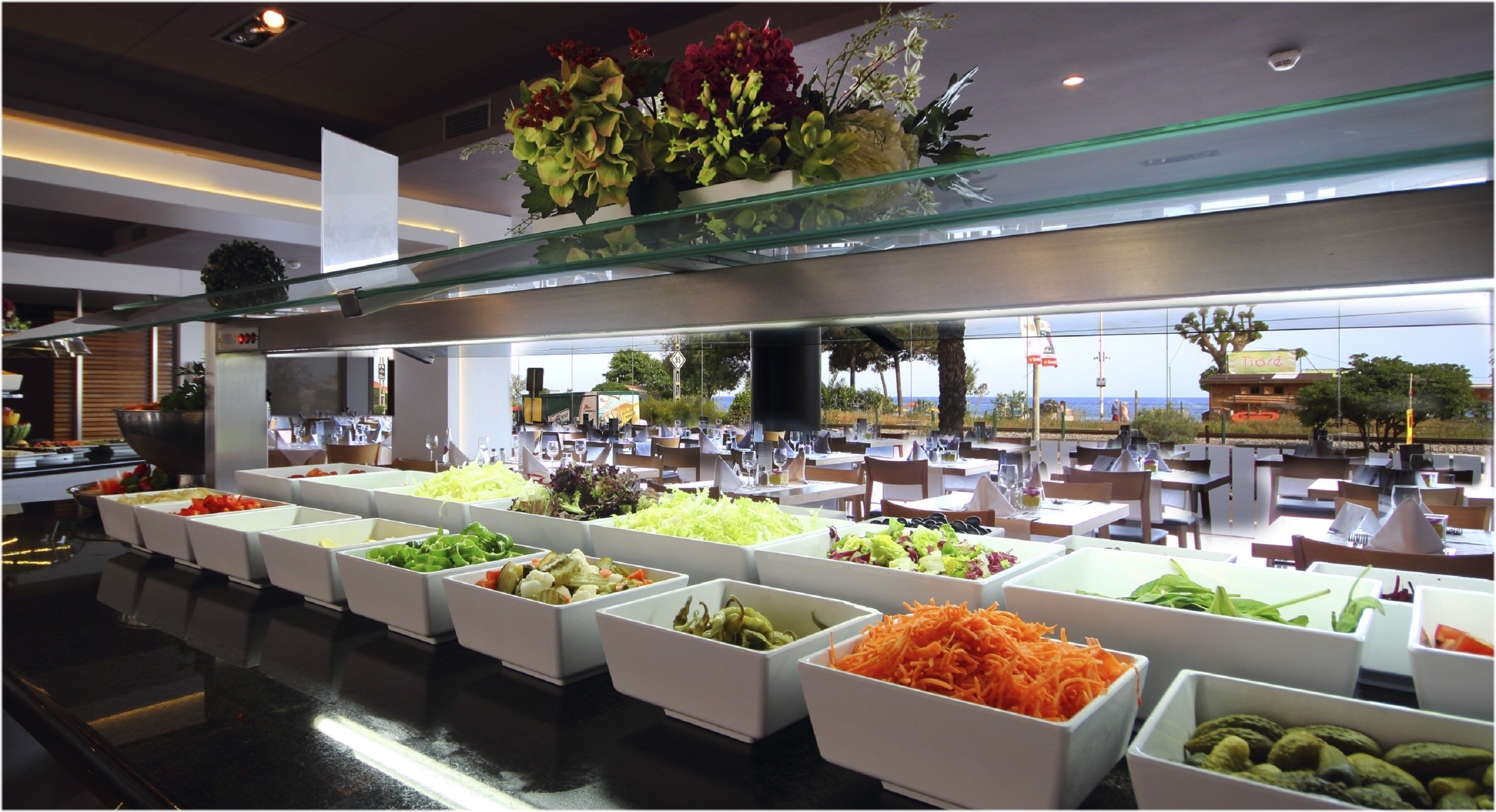 Buffet com diversas opções de comida em tigelas brancas, prateleira de vidro com flores e vista para a área de estar externa.