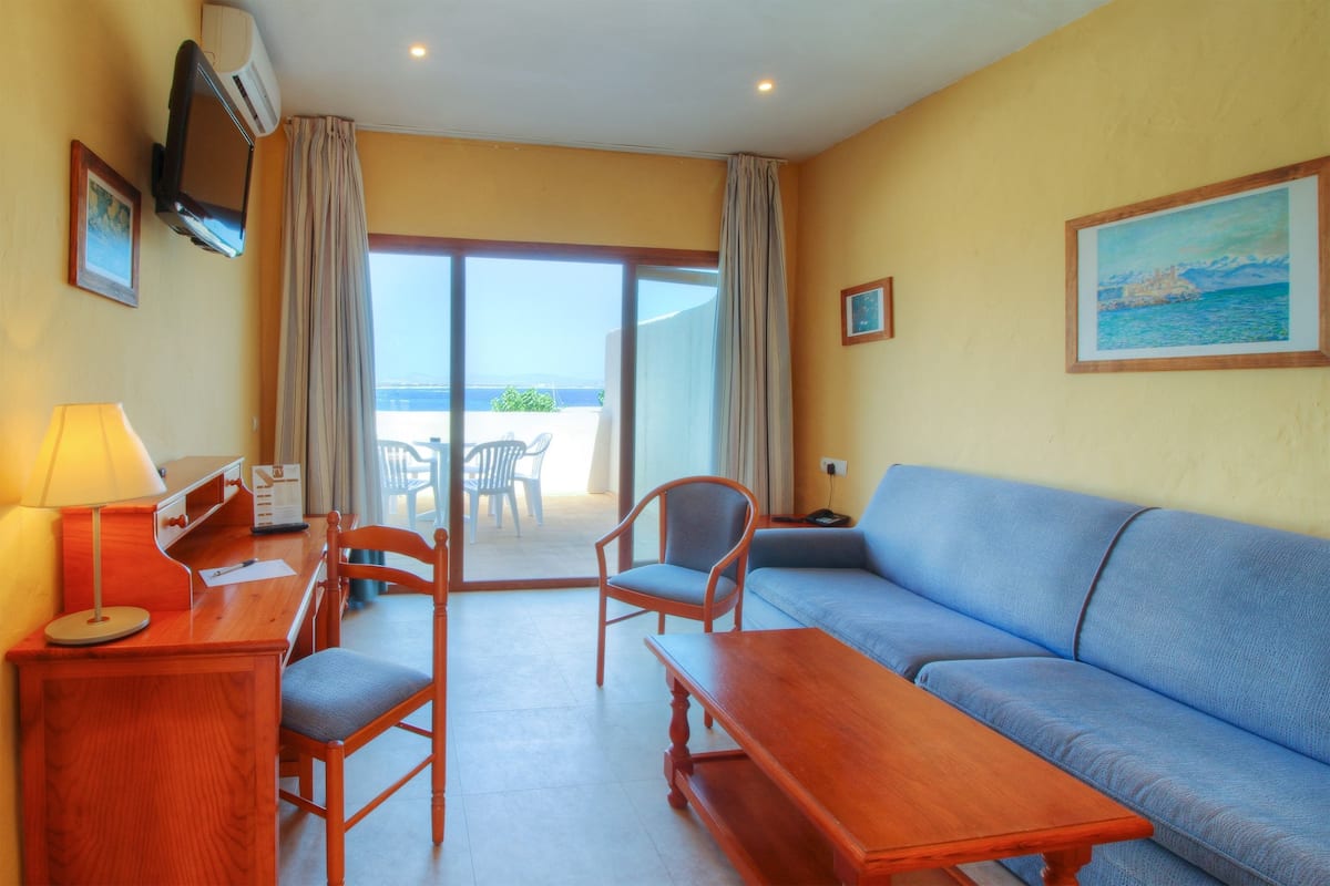 Quarto com sofá azul, mesa de madeira e cadeiras, escrivaninha com luminária e um quadro na parede.