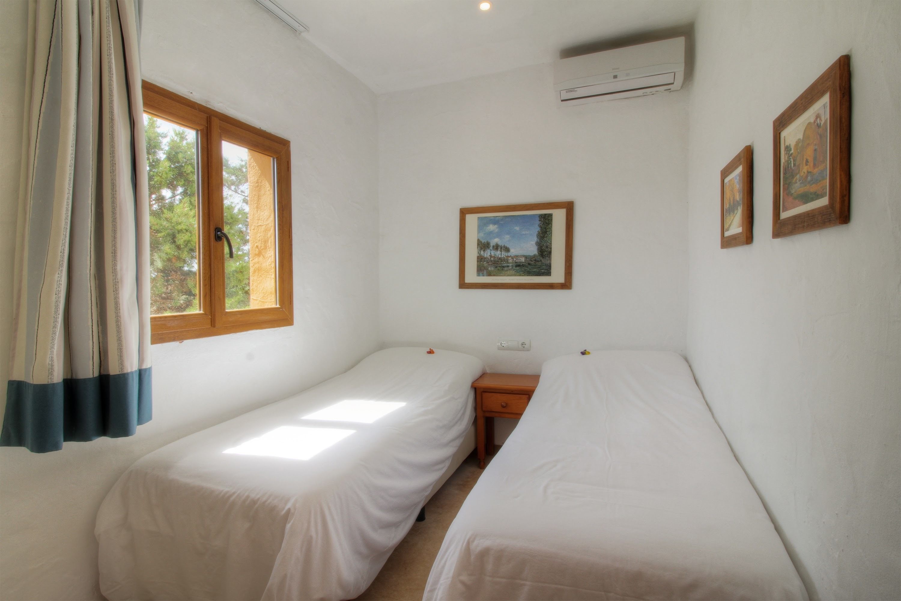 Quarto com duas camas individuais, uma mesinha de cabeceira de madeira, uma janela com cortinas e dois quadros na parede.