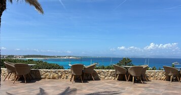 Terraço com cadeiras de vime e muro de pedra, com vista para a costa onde se vêem barcos e edifícios.