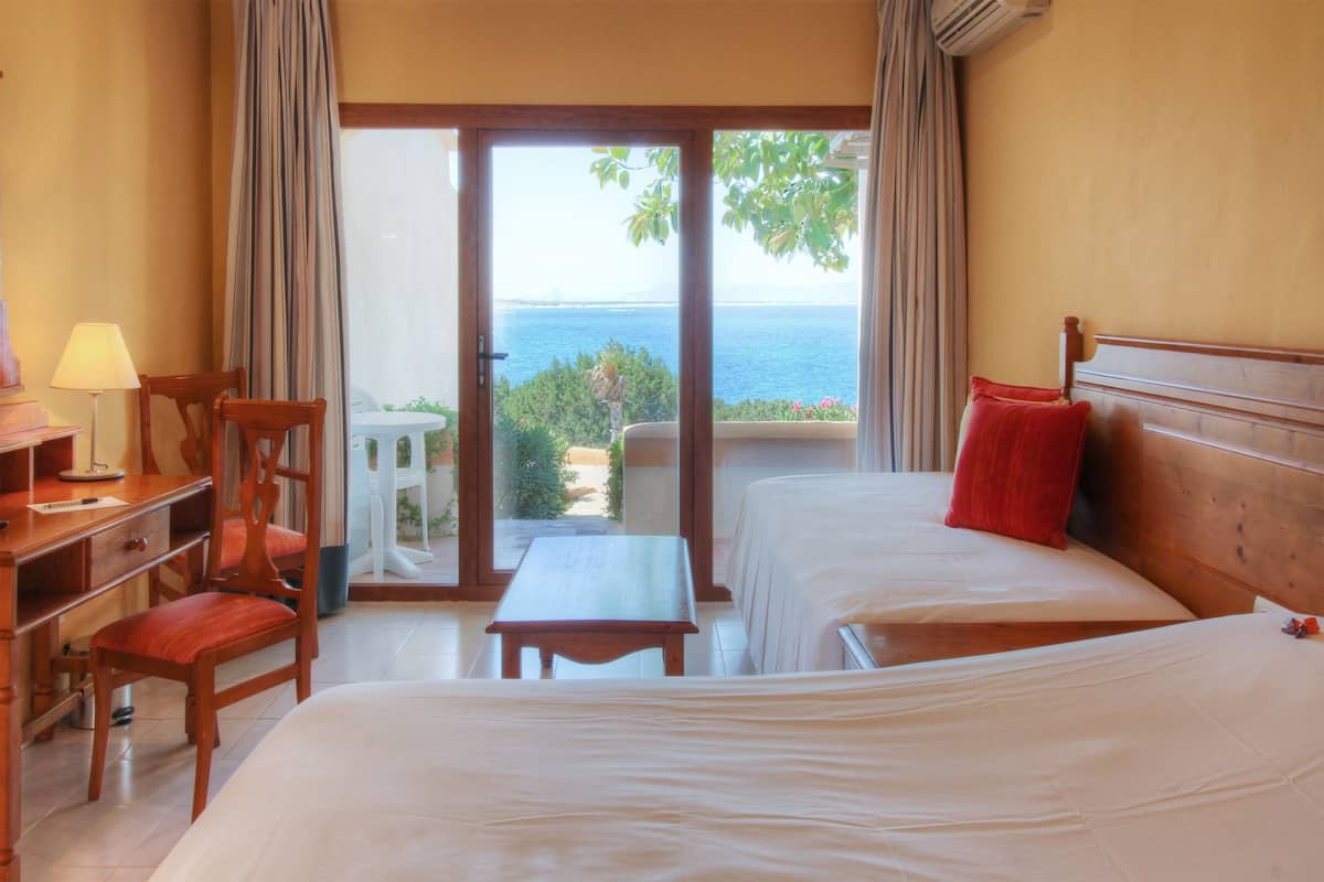 Quarto de hotel com duas camas, uma secretária e uma cadeira. Vê-se o mar através de uma porta deslizante.