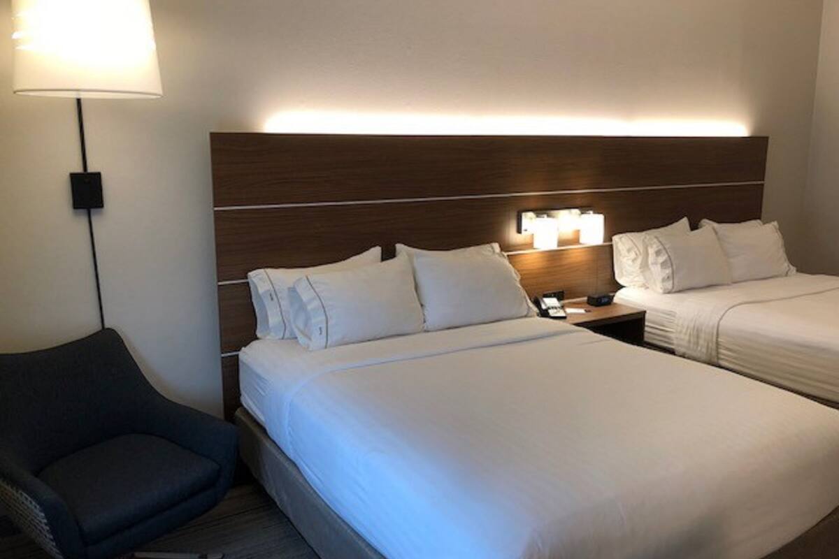 Quarto de hotel com duas camas, uma mesa de cabeceira, uma luminária e uma cadeira.