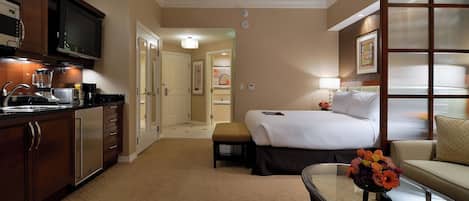 Quarto de hotel com cozinha compacta, uma cama, um sofá e uma mesa de centro de vidro com flores.