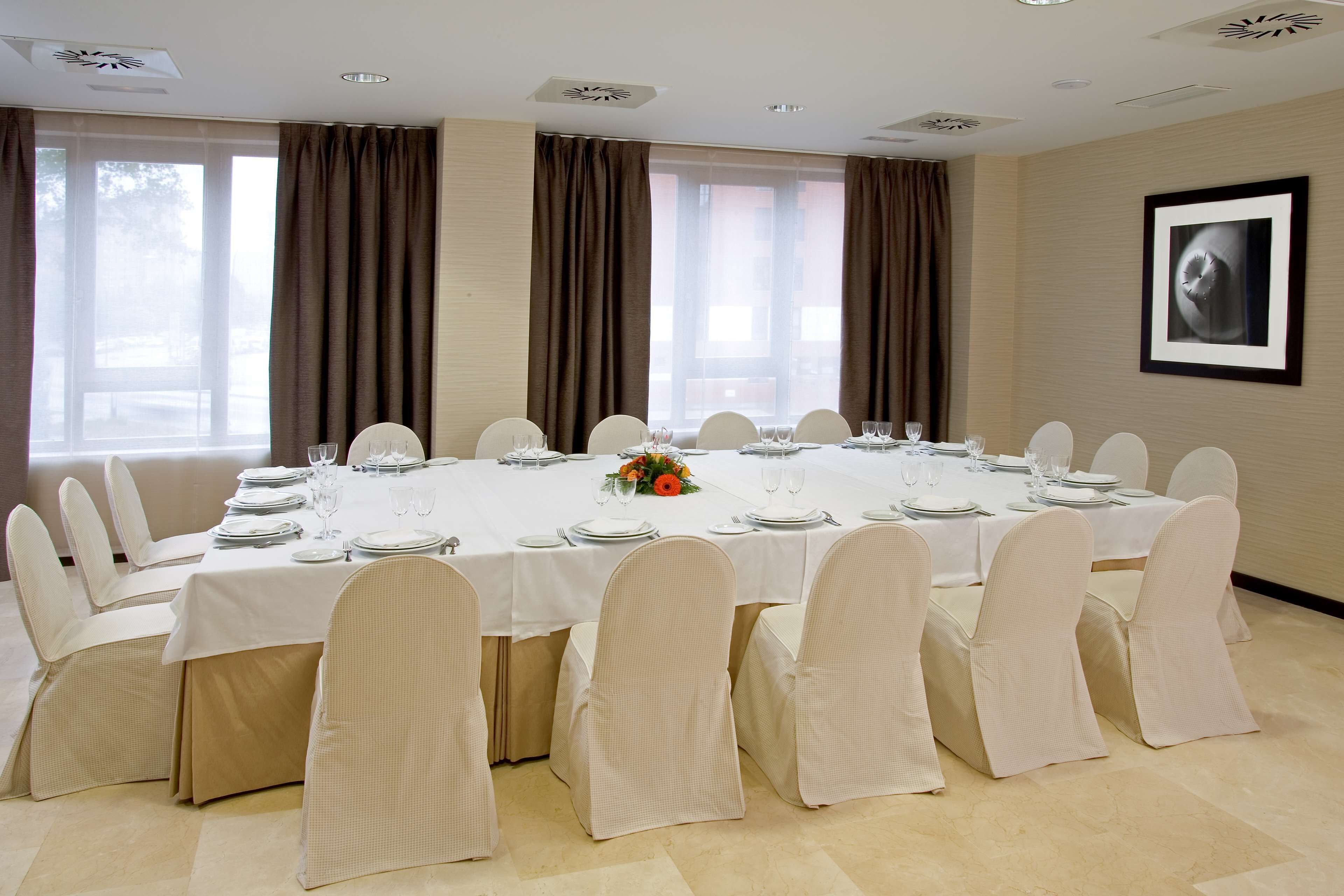 Uma sala de conferência com uma mesa redonda posta para uma reunião, cadeiras bege e um quadro na parede.