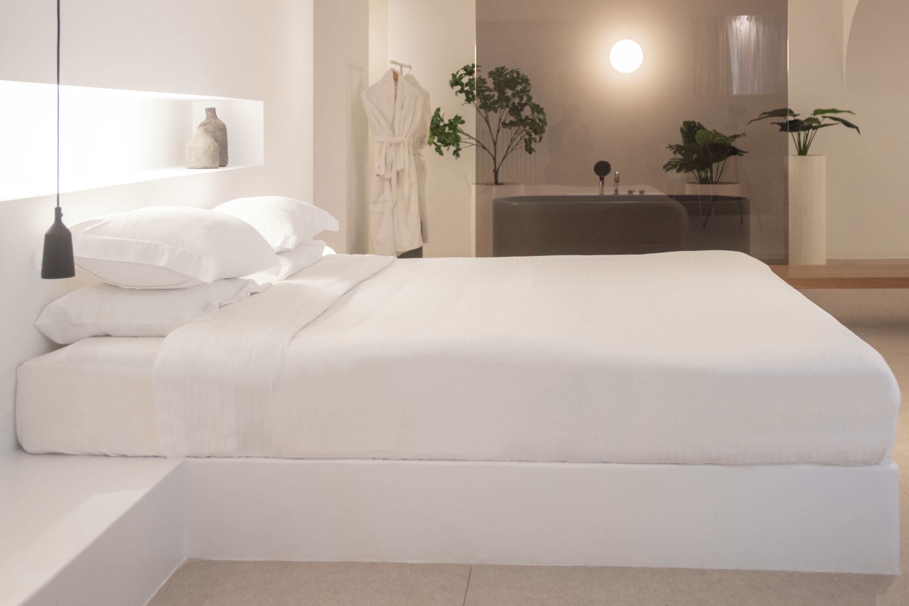 Quarto moderno com cama grande, mesas de cabeceira, cabideiro suspenso, planta e espelho redondo.