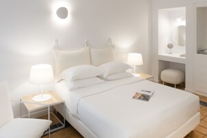 Quarto de hotel com cama bem arrumada, duas mesas de cabeceira com luminárias, um espelho redondo pequeno e uma cadeira.