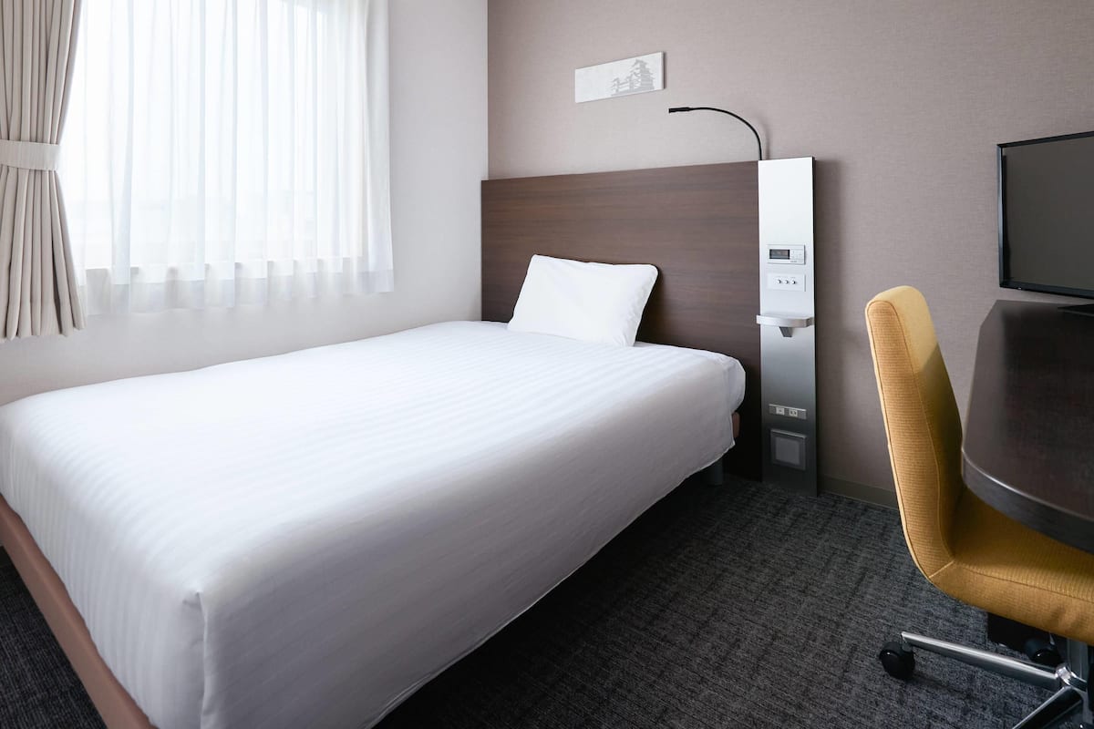 Quarto de hotel com uma cama grande, uma escrivaninha com cadeira, televisão e painel de controle fixado na parede.