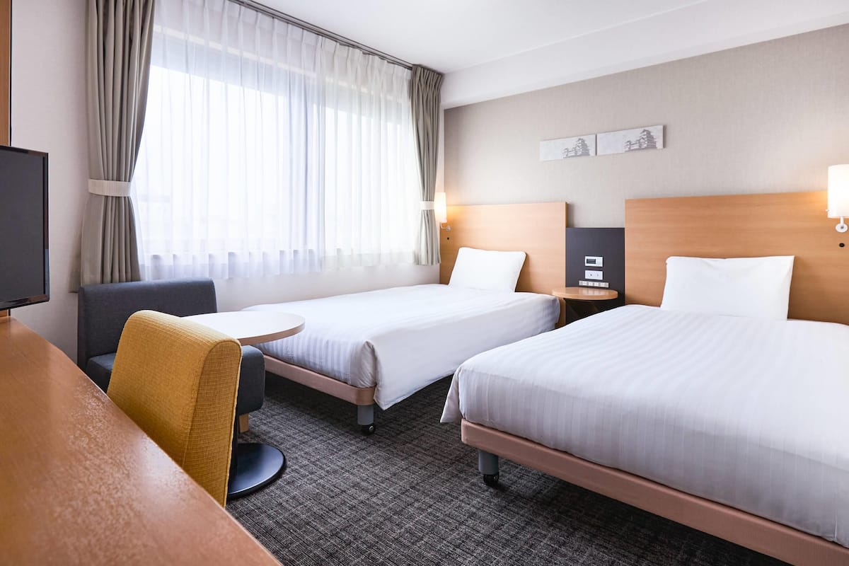 Quarto de hotel com duas camas, uma escrivaninha, uma cadeira, uma mesinha e uma televisão.