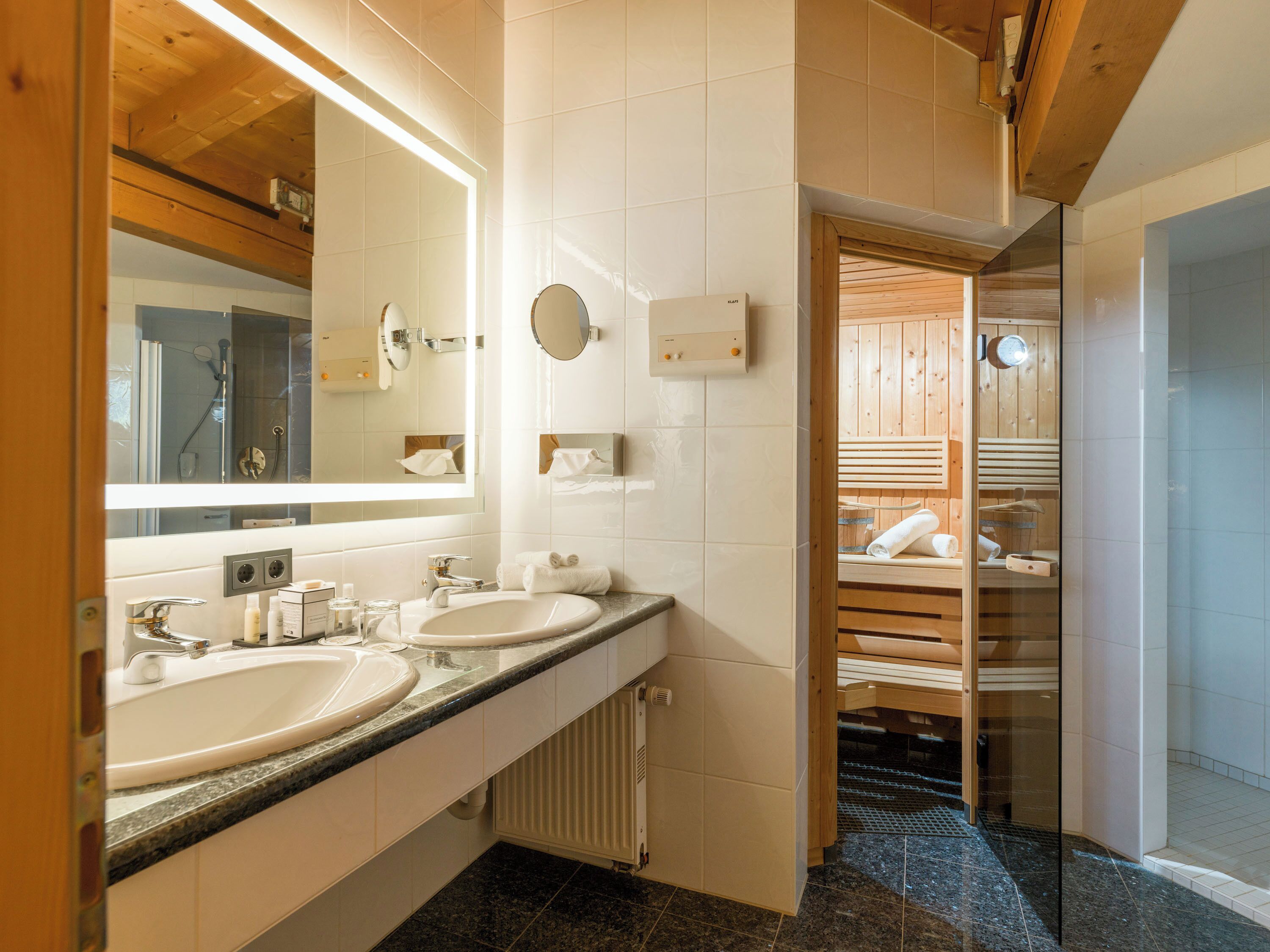 Casa de banho moderna com duas bacias, um espelho grande e uma sauna de madeira visível através de uma porta aberta.