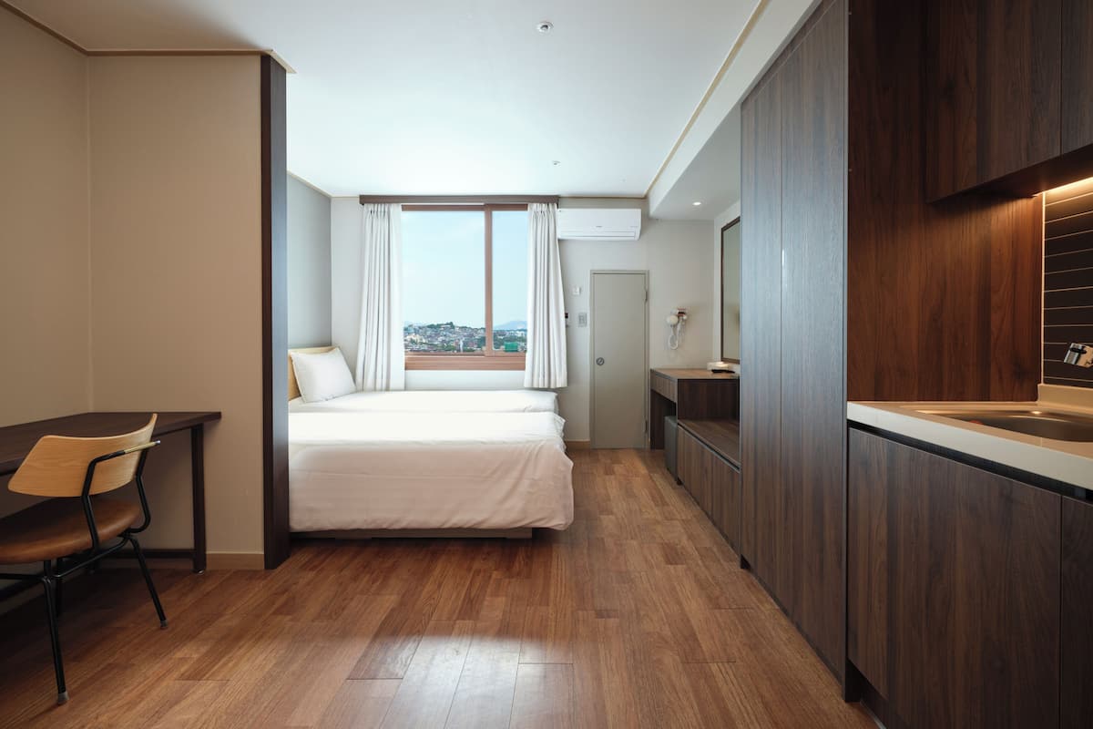 Quarto de hotel moderno com cama, escrivaninha, cadeira, pequena cozinha e vista para a cidade através de grandes janelas.