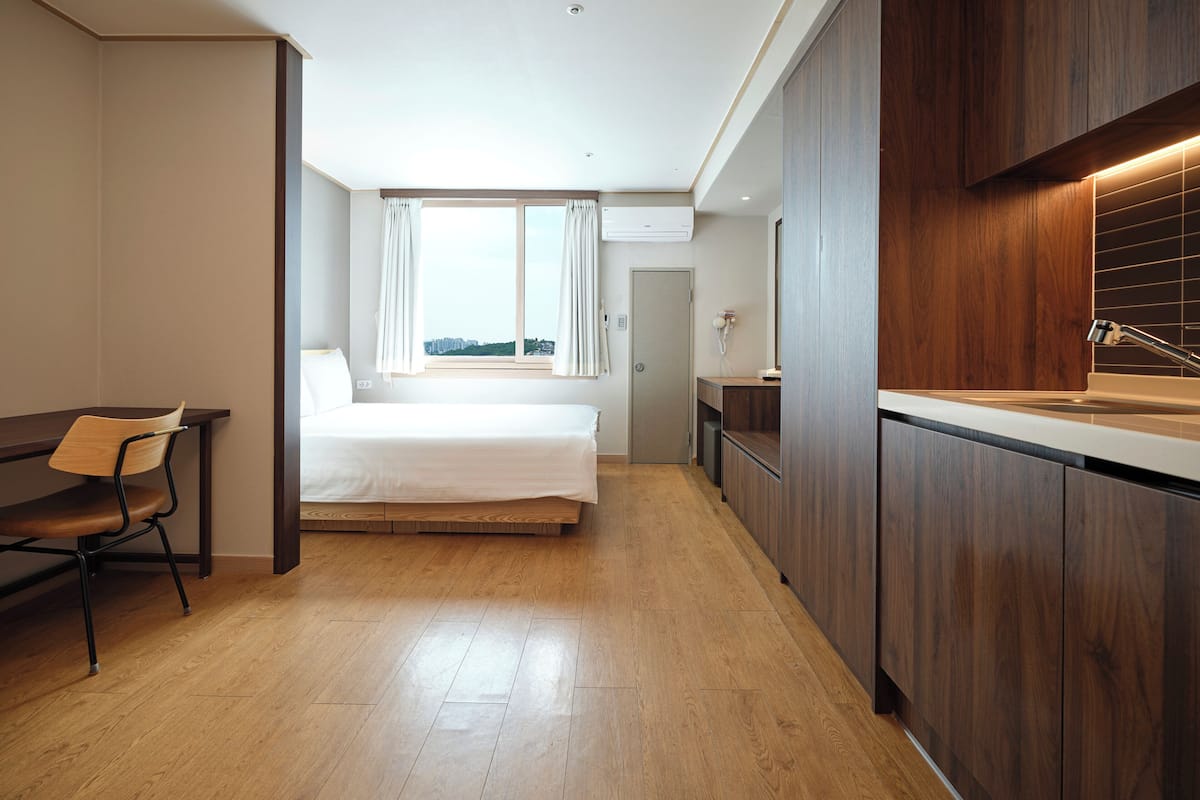 Quarto de hotel moderno com cama, escrivaninha, janela com cortinas, área de cozinha e vista para a paisagem urbana.