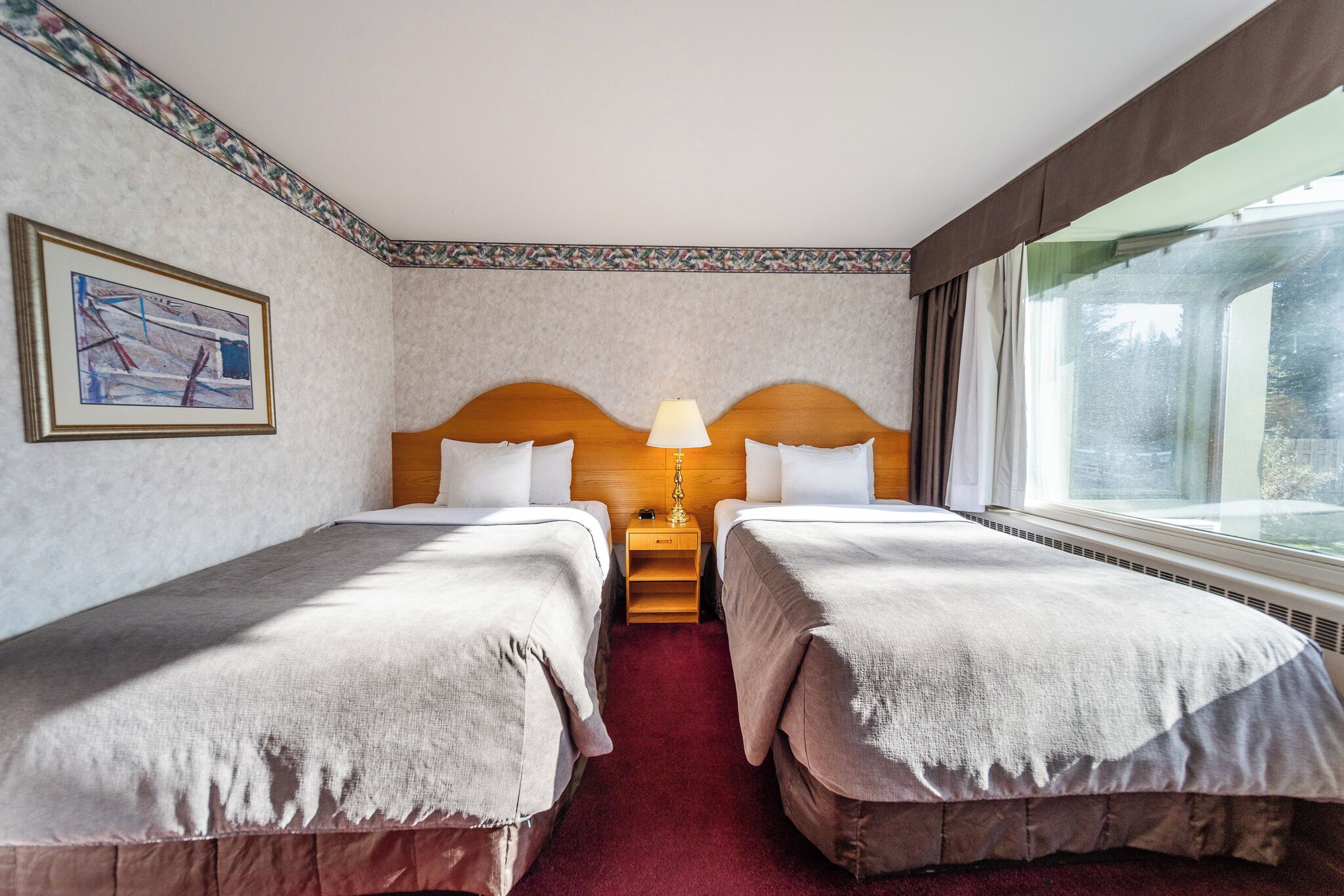 Quarto de hotel com duas camas, cabeceira de madeira, mesa de cabeceira, abajur, quadro emoldurado e uma janela grande.