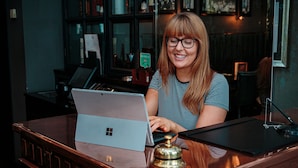 Mulher no balcão de recepção com um laptop, uma mesa de madeira e um sino de latão.