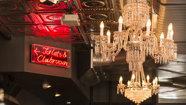 Uma área de bar com superfície refletora, um candelabro e uma placa de néon indicando sanitários e salão de festas.