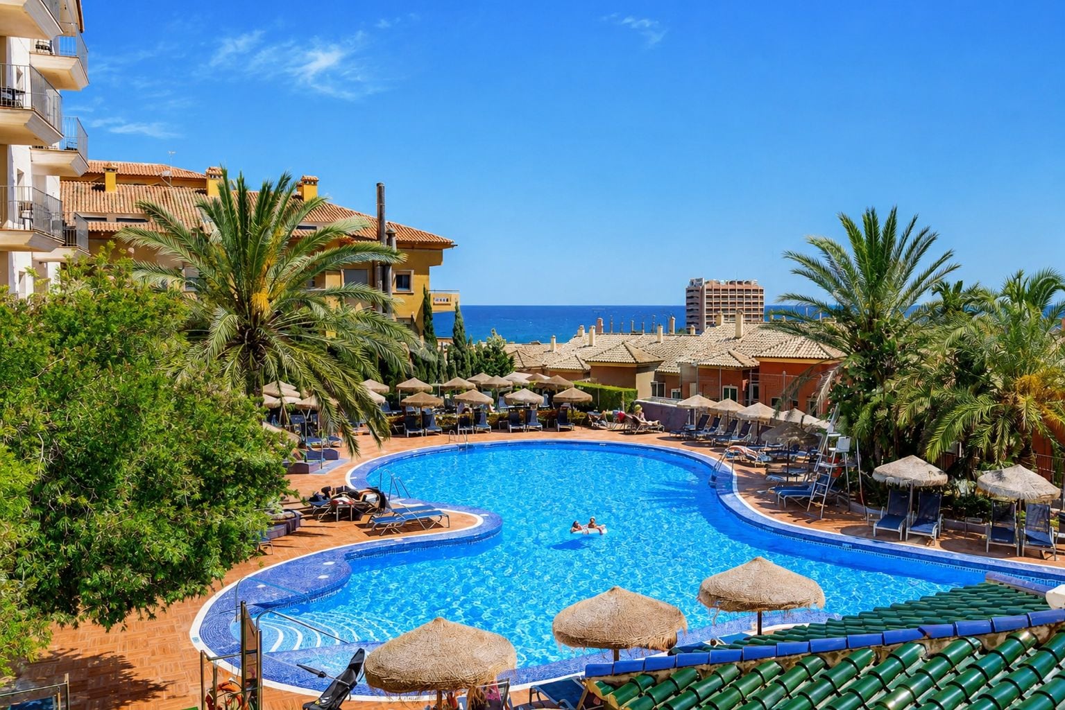 Piscina de resort com design curvo, cercada por espreguiçadeiras e guarda-sóis, com vista para o mar e edifícios ao fundo.
