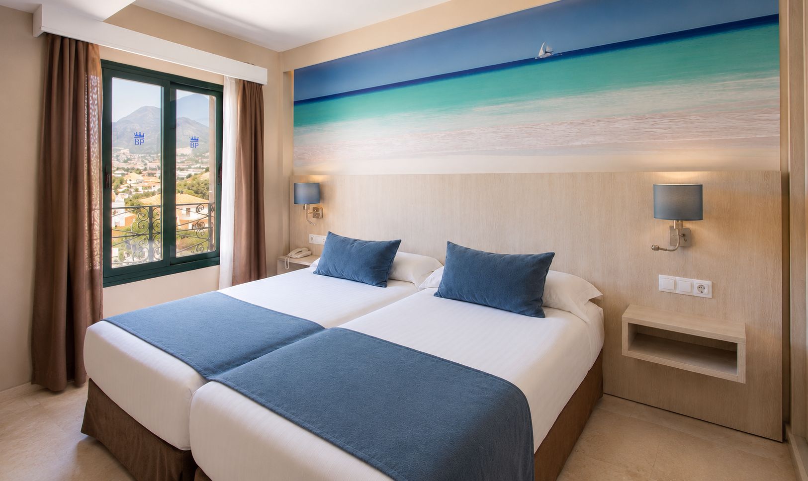 Quarto de hotel com uma cama grande, duas mesas de cabeceira, vista para o mar e um quadro de uma cena praiana.
