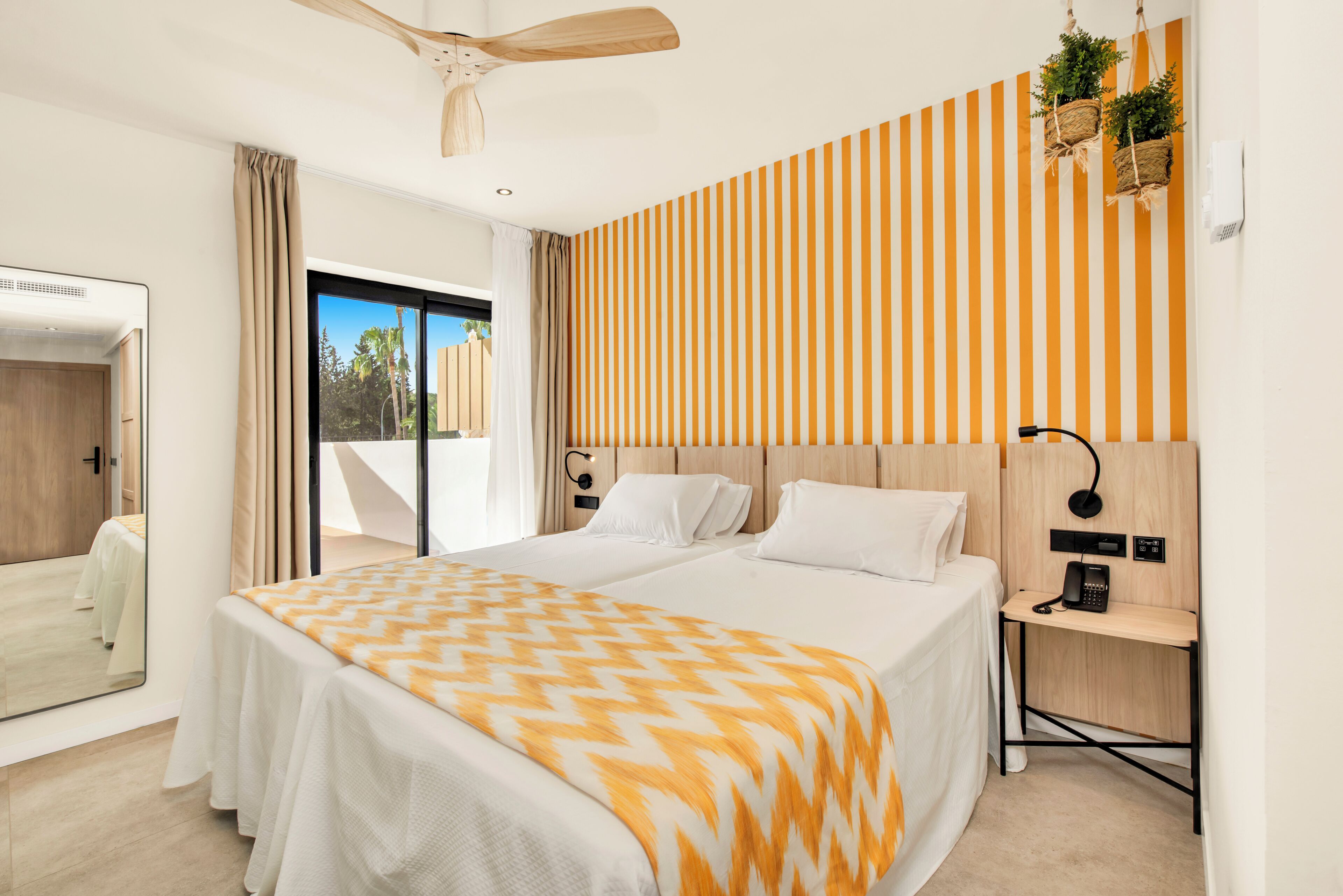 Quarto de hotel com cama, escrivaninha com telefone, ventoinha no teto e vista através de uma porta de vidro.